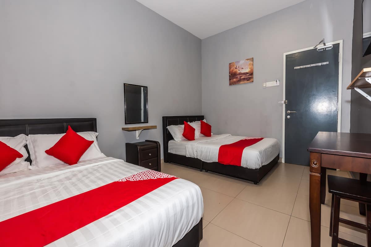 Quarto de hotel com duas camas, cada uma com roupa de cama branca e detalhes vermelhos, pequenas mesas de cabeceira, prateleira na parede, uma porta e uma escrivaninha com cadeira.