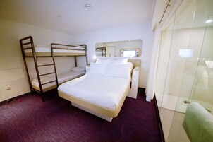 Quarto de hotel com cama beliche, uma cama de solteiro, mesa de cabeceira com abajur, espelho e box de chuveiro em vidro.