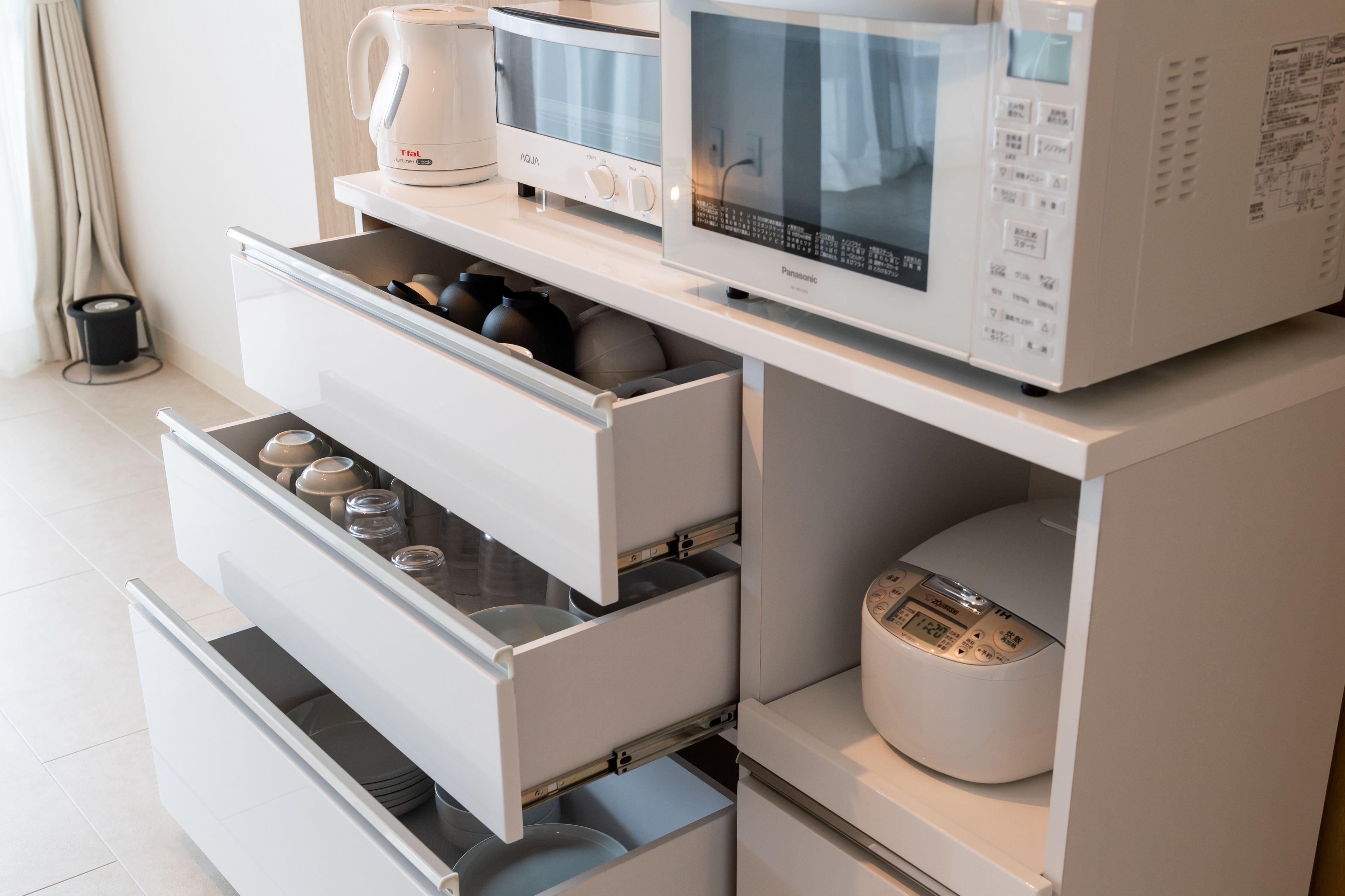 Cozinha equipada com micro-ondas, torradeira e cafeteira na bancada. Existem gavetas e uma geladeira abaixo.