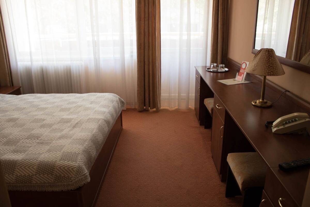 Quarto de hotel com uma cama, uma escrivaninha com abajur, um telefone e uma pequena mesa com um copo.