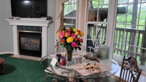 Uma sala de estar com lareira, televisão, mesa de jantar com um vaso de flores e vista para um deck de madeira através de grandes janelas.
