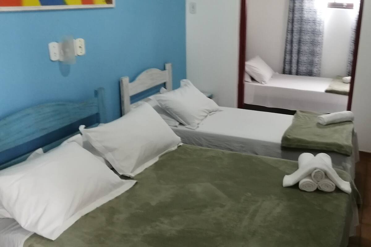 Quarto com duas camas, uma com colcha verde e almofadas brancas, e outra com colcha branca e almofadas brancas. Há uma mesinha com um telefone e um objeto decorativo. As paredes do quarto são azuis e há uma janela com cortina.