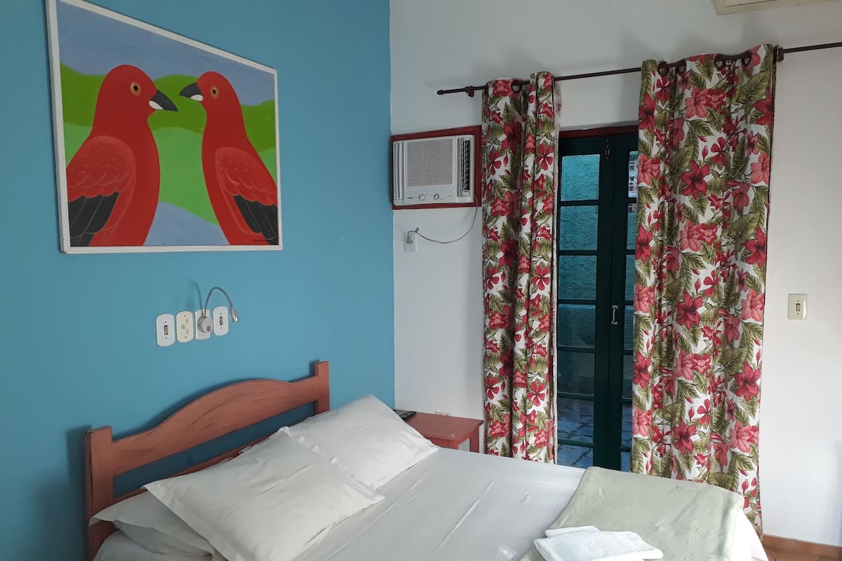 Quarto com cama, ar-condicionado fixado na parede, tomada e janela com cortinas florais.