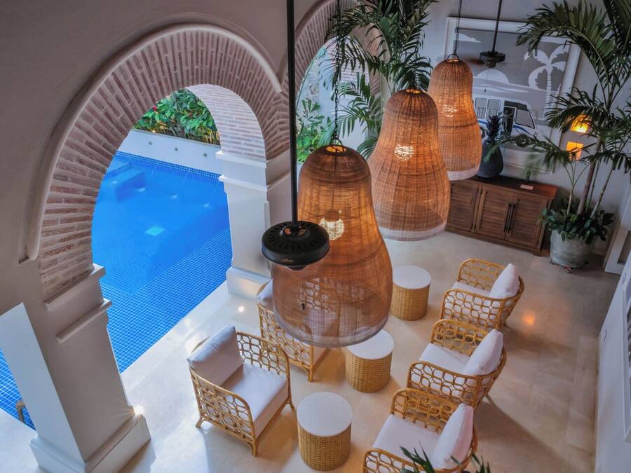 Área de piscina interior luxuosa, com vista para uma piscina azul, mobiliário de vime e luminárias pendentes.