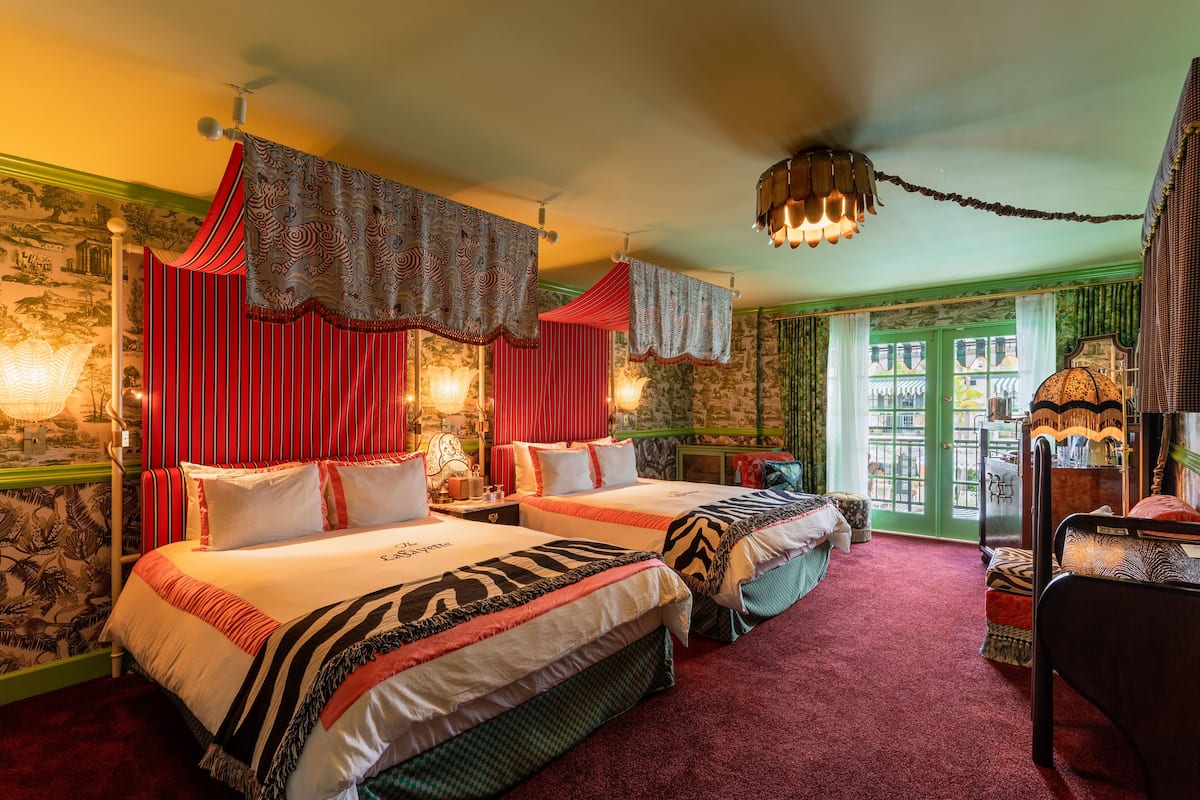 Quarto com duas camas, cortinas vermelhas, roupa de cama estampada, um lustre e vista através de uma porta verde.