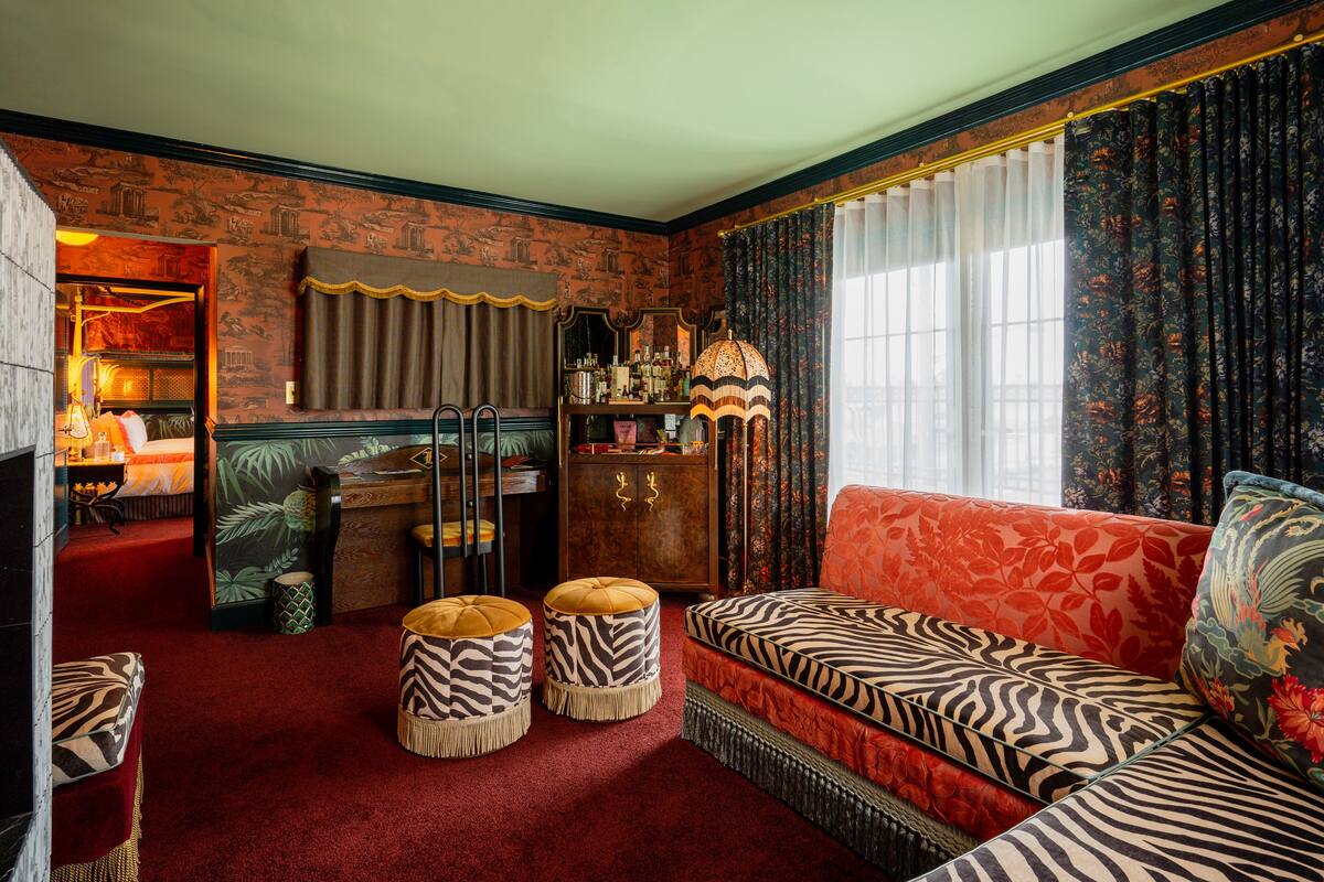 Quarto com tapete vermelho, sofá estampado com padrão de zebra, dois pufes, um armário de madeira e uma janela grande com cortinas.