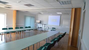 Uma sala de conferências com uma mesa comprida, cadeiras verdes, uma tela de projetor e um quadro branco.