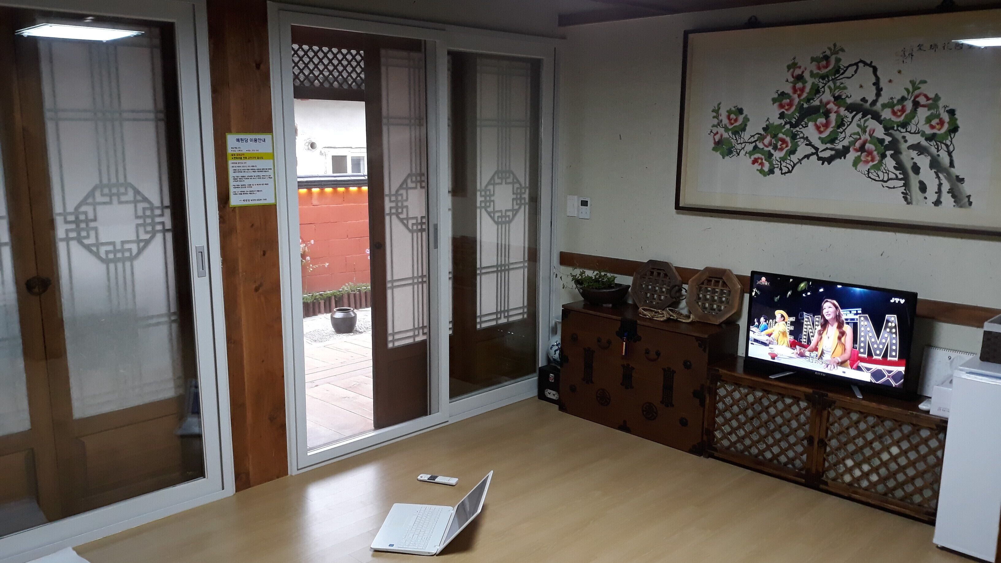 Quarto com piso de madeira, uma TV em um móvel, um laptop no chão e uma porta com um painel decorativo.