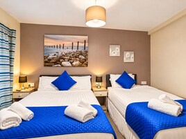 Quarto de hotel com duas camas, cada uma com colcha azul acolchoada e roupa de cama branca, um quadro com um pôr do sol sobre rochas na parede e uma luminária sobre as camas.