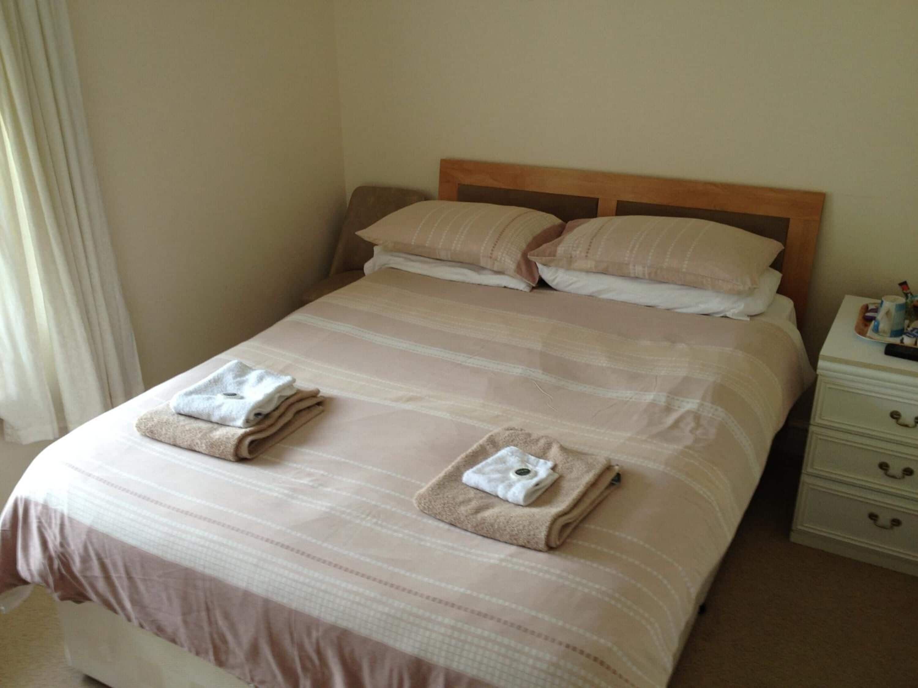 Cama bem arrumada com duas toalhas dobradas, cabeceira de madeira e mesa de cabeceira branca com gavetas.