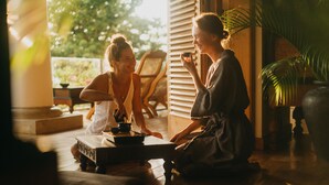 Duas pessoas sentadas no chão, uma segurando uma xícara, em um quarto com móveis de madeira e uma mesa com um bule.