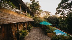 Uma trilha de pedra leva a uma área de piscina com um edifício de telhado de palha e vegetação exuberante.