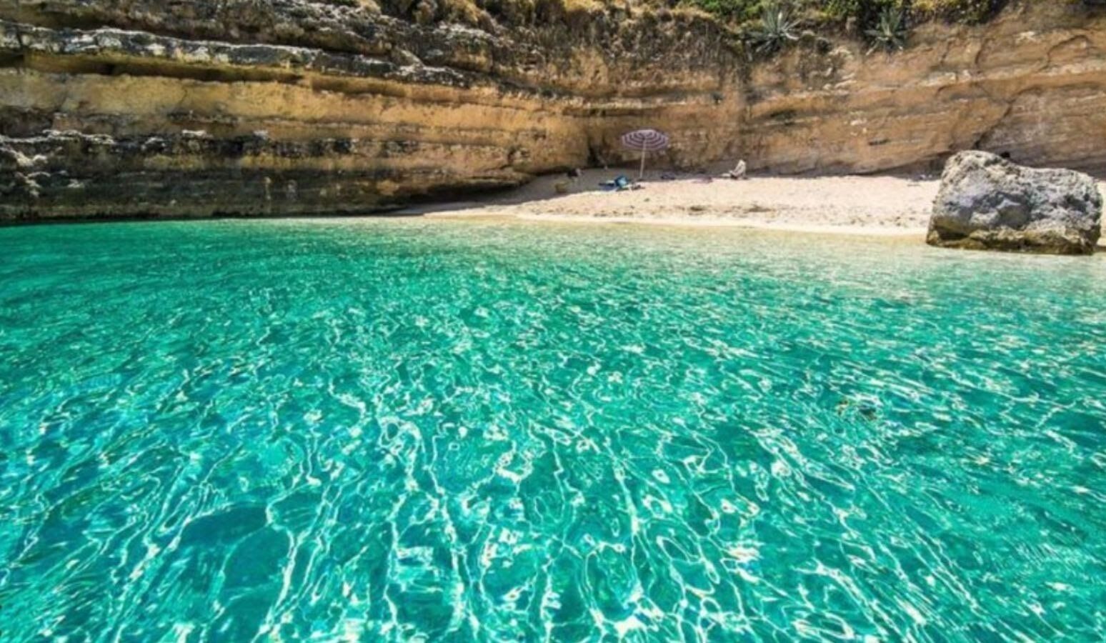 Mar azul-turquesa límpido, com um penhasco rochoso e uma praia arenosa ao fundo.