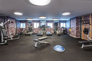 Uma academia com diversos equipamentos de exercício, paredes com padrão de tijolos e grandes janelas.