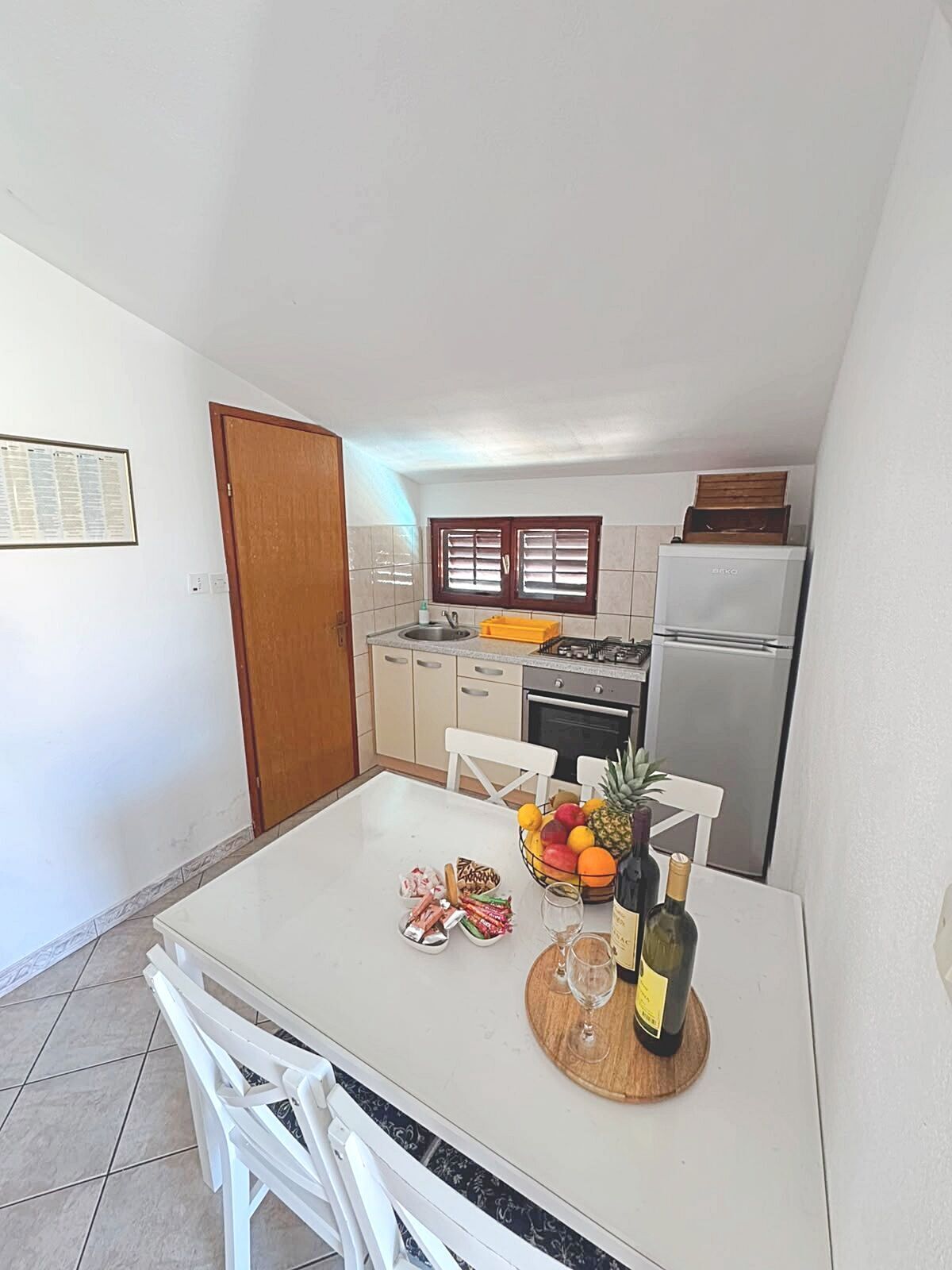 Uma cozinha compacta com mesa, cadeiras, um cesto de frutas e uma garrafa de vinho.
