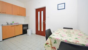 Cozinha compacta com armários de madeira, uma pia e um fogão. Há uma mesa de jantar com toalha de mesa florida e duas cadeiras. Uma lixeira branca está visível perto da porta.
