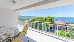 Varanda com mesa e cadeiras, guarda-corpo branco e vista para o mar e colinas.
