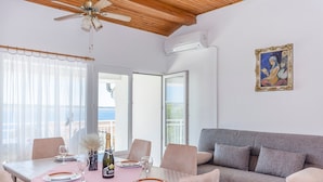 Uma área de refeições com uma mesa posta para dois, um sofá cinzento e vista para o mar através de grandes janelas.