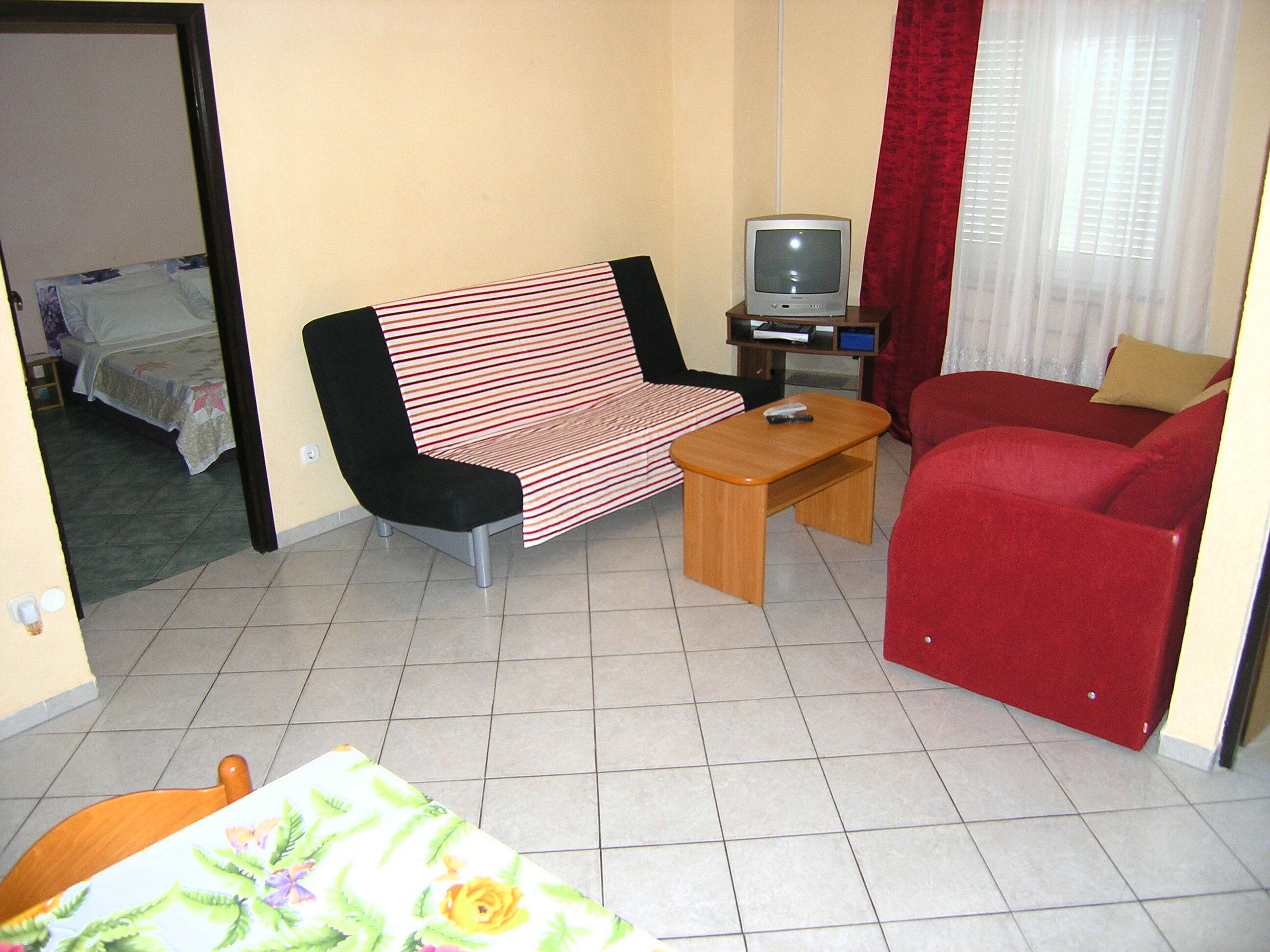 Uma sala de estar pequena com um sofá listrado, uma poltrona vermelha, uma mesa de centro de madeira e uma televisão sobre um móvel.