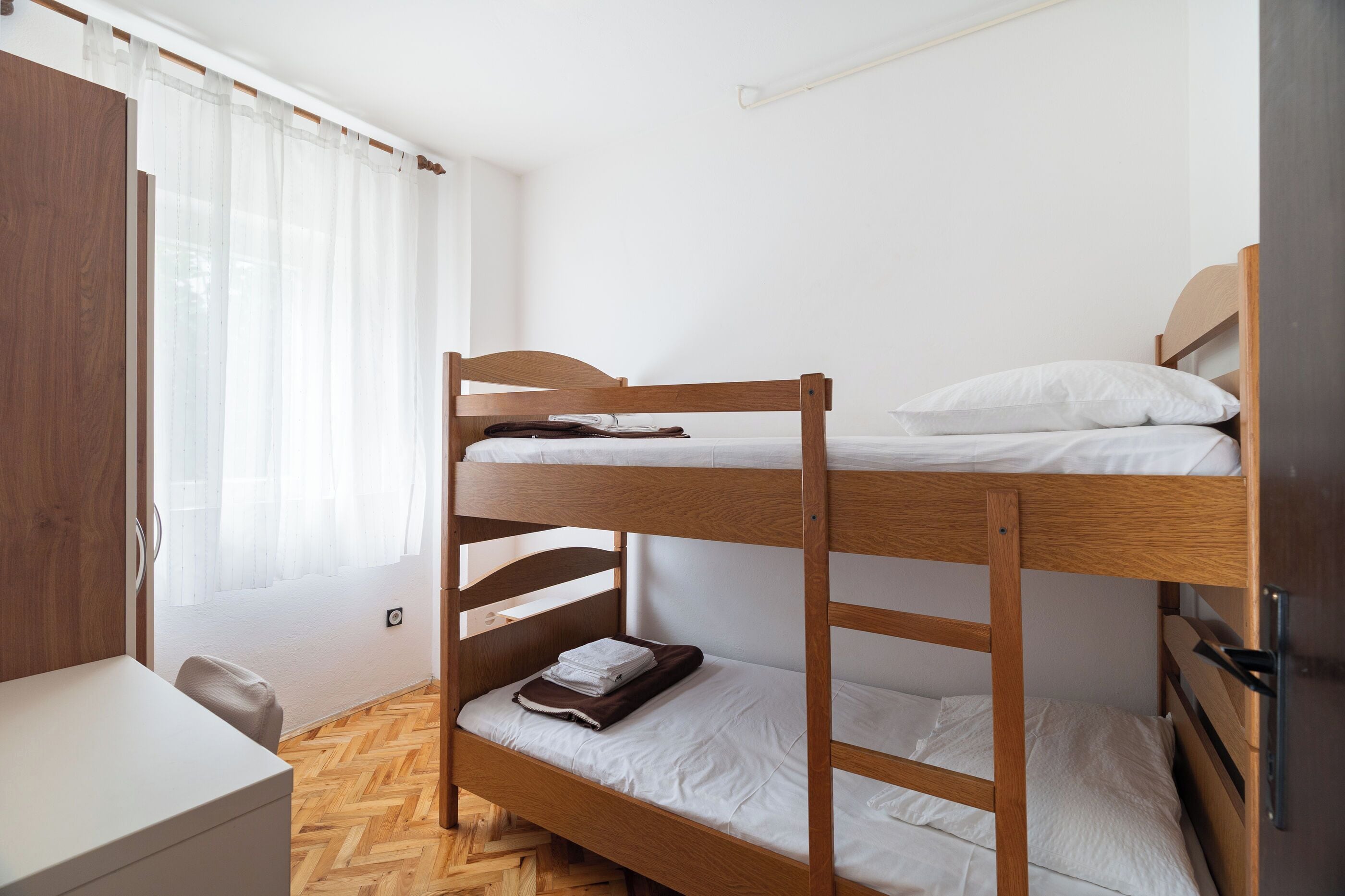 Uma cama de solteiro com escrivaninha e cadeira, um guarda-roupa e uma janela com cortinas.
