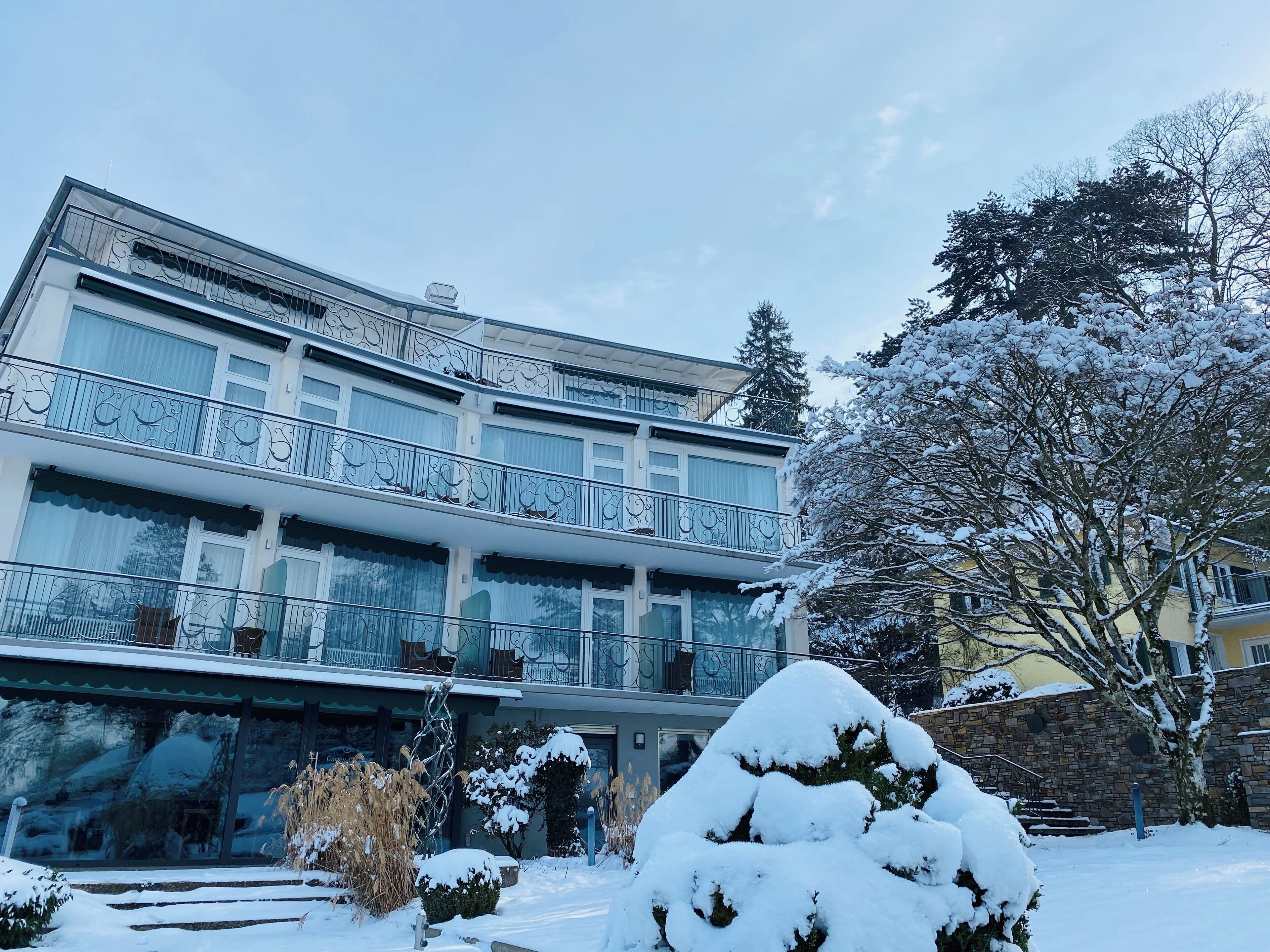 Um hotel com várias andares, varandas, um jardim coberto de neve e um muro de pedra.