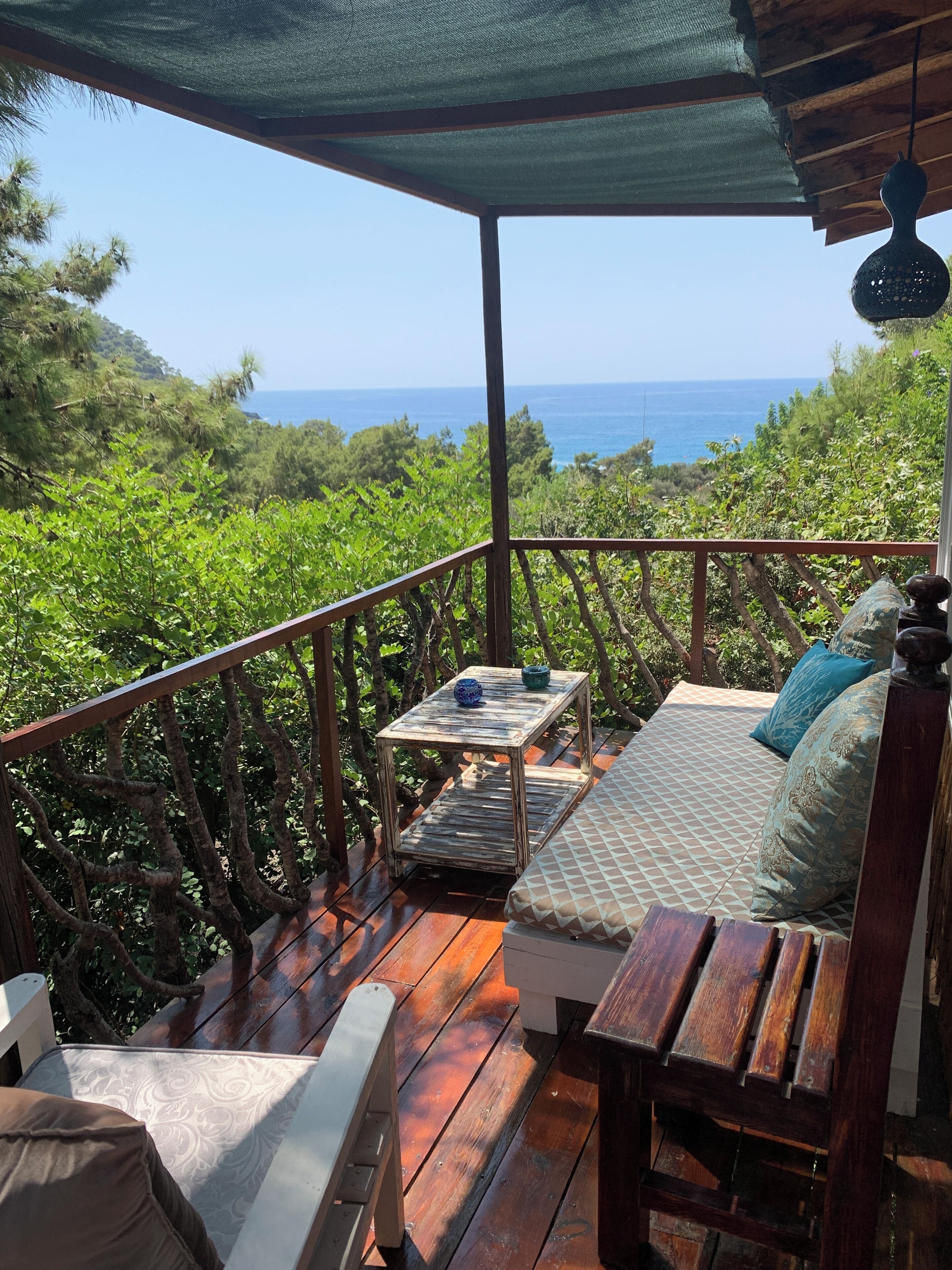 Um deck de madeira com sofá e mesa, com vista para uma paisagem exuberante e o mar.