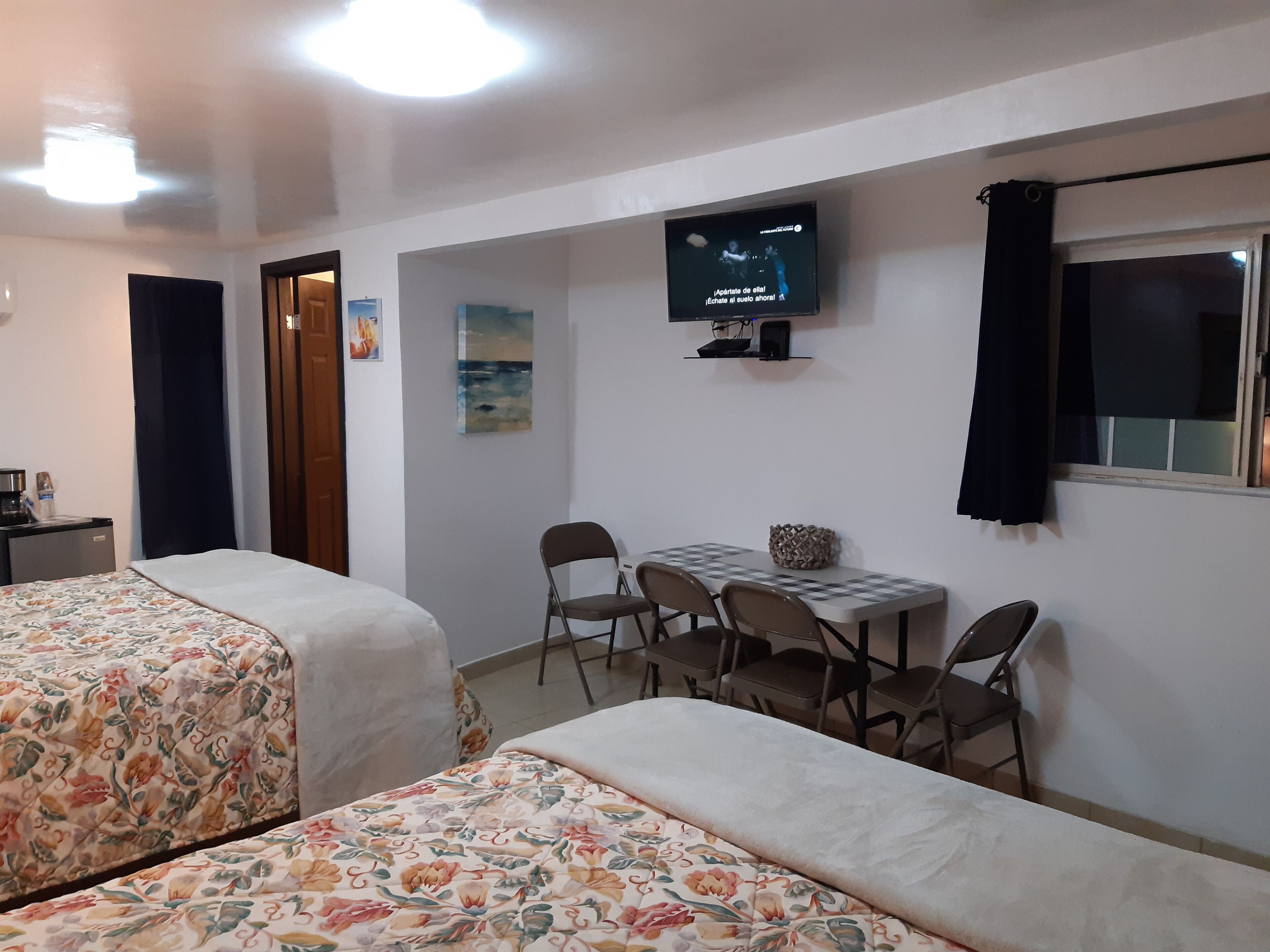 Quarto de hotel com duas camas, uma mesinha com cadeiras, TV fixada na parede e janela com cortinas.