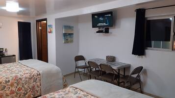Quarto de hotel com duas camas, uma mesinha com cadeiras, TV fixada na parede e janela com cortinas.
