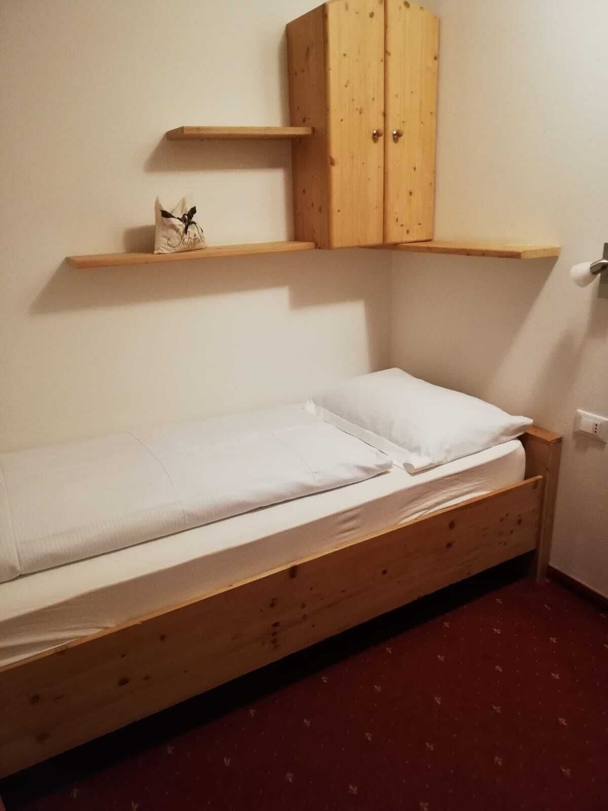 Quarto pequeno com uma cama de solteiro, estrutura de madeira, guarda-roupa de madeira e uma pequena prateleira sobre a cama.