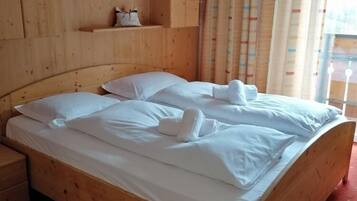 Uma cama de madeira com lençóis e toalhas brancas, um criado-mudo de madeira, uma estante com uma bolsa e uma janela com cortinas.