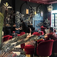 Um restaurante com cadeiras estofadas vermelhas, decoração na parede preta e dourada e grandes vasos de plantas.