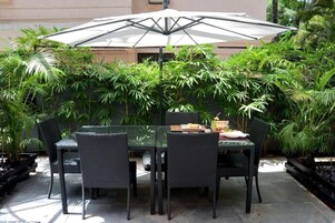 Mesa com tampo de vidro, quatro cadeiras e um grande guarda-sol em ambiente externo com vegetação exuberante.