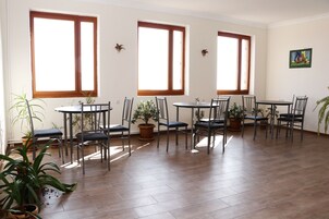 Uma ampla sala de jantar com várias mesas e cadeiras, janelas grandes e plantas em vasos.