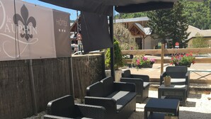 Uma área externa com mesas e cadeiras de vime sob uma cobertura, uma placa com 'le AUPHIN HOTEL' e vista para a área circundante, incluindo edifícios e vegetação.