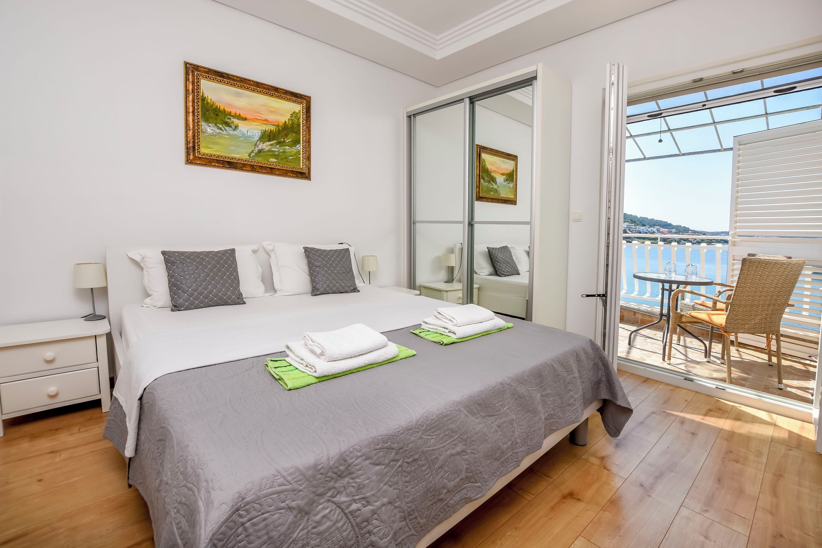 Quarto com cama, mesa de cabeceira, penteadeira, varanda com cadeira e um quadro na parede.