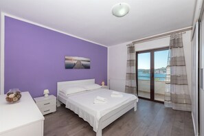 Quarto com cama, mesa de cabeceira, luminária, vaso com objetos decorativos e vista através de uma porta de vidro.