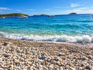Uma praia de pedras com águas azul-claras e barcos ao longe.