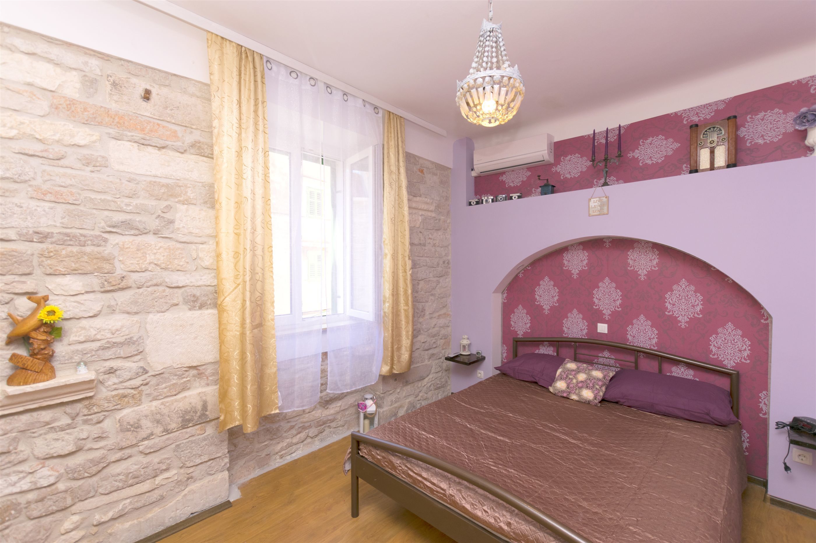 Quarto com parede de pedra, cama com cabeceira, candelabro pendurado e janela com cortinas finas.