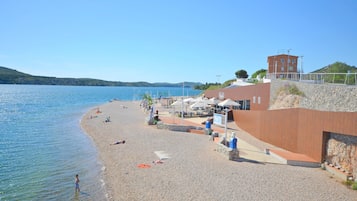 Uma praia com costa de pedras, um edifício com fachada vermelha e um céu azul e límpido.