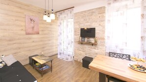Uma sala de estar moderna com piso de madeira, um sofá preto, uma mesa de centro de madeira, uma TV fixada em uma parede de pedra e um espaço de jantar com mesa e cadeiras de madeira.