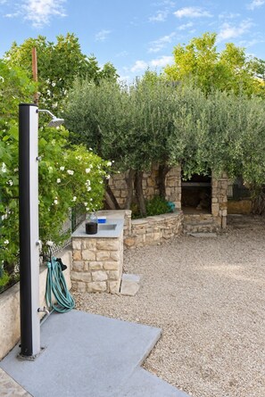Área externa em pedra com um banco de pedra, estrutura metálica e um jardim com oliveiras.