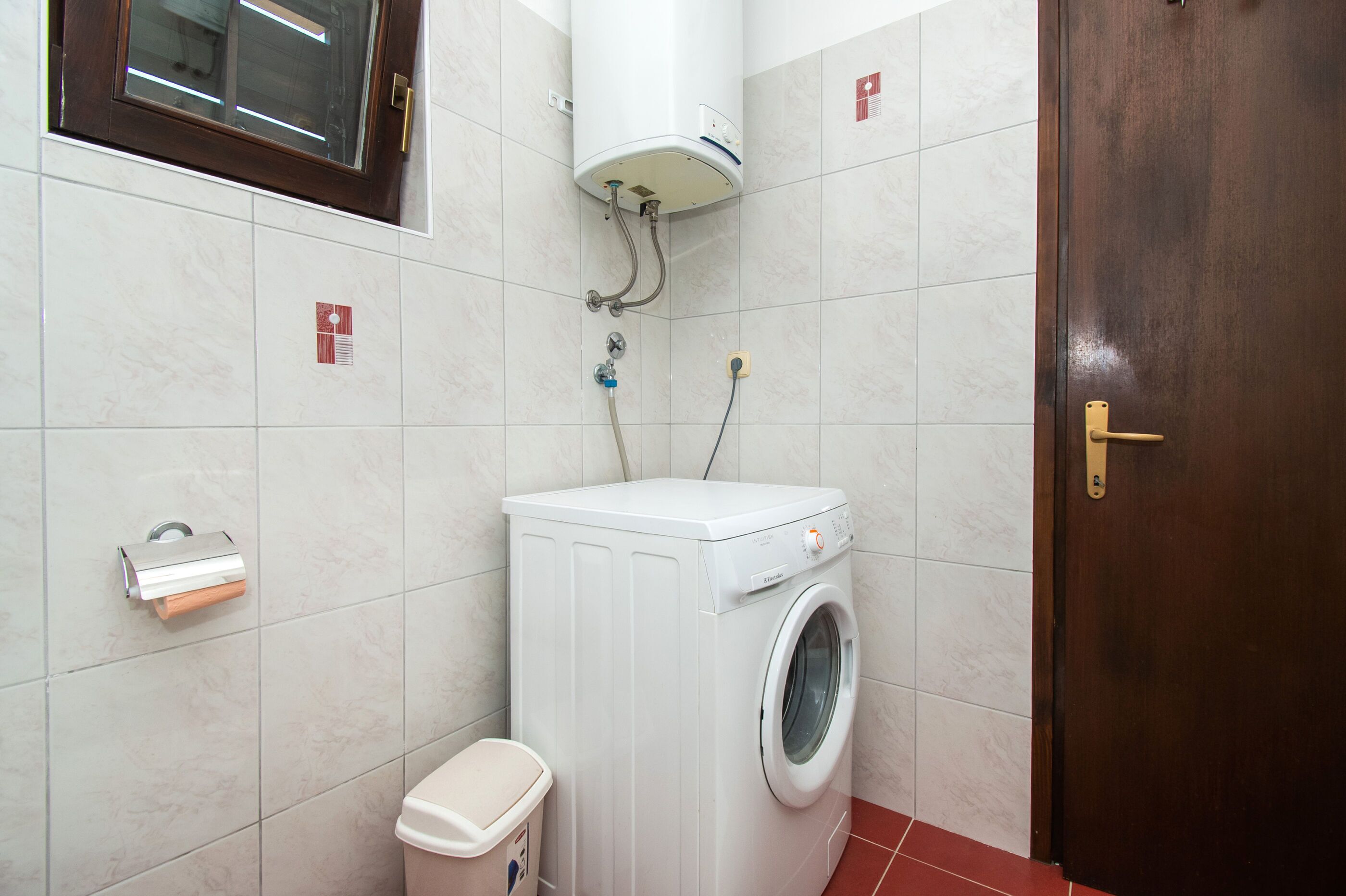 Uma lavanderia compacta com máquina de lavar, uma pequena lixeira e aquecedor de água na parede.