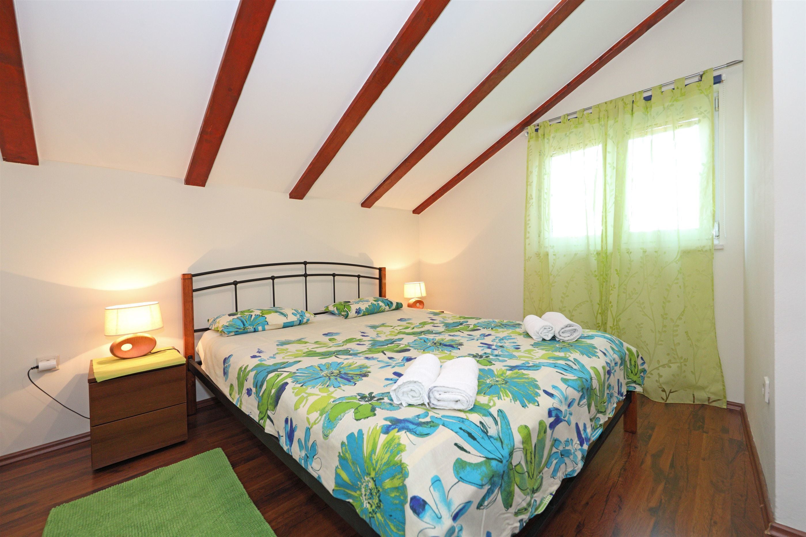 Quarto com cama, mesa de cabeceira, abajur e janela com cortinas.