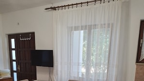 Uma sala de jantar com uma mesa retangular e seis cadeiras, uma televisão sobre um móvel, e uma porta com cortina.