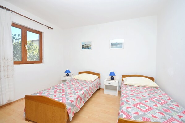Quarto com duas camas de solteiro, cada uma com uma mesa de cabeceira e uma luminária. Há uma janela com cortinas e duas imagens emolduradas na parede.