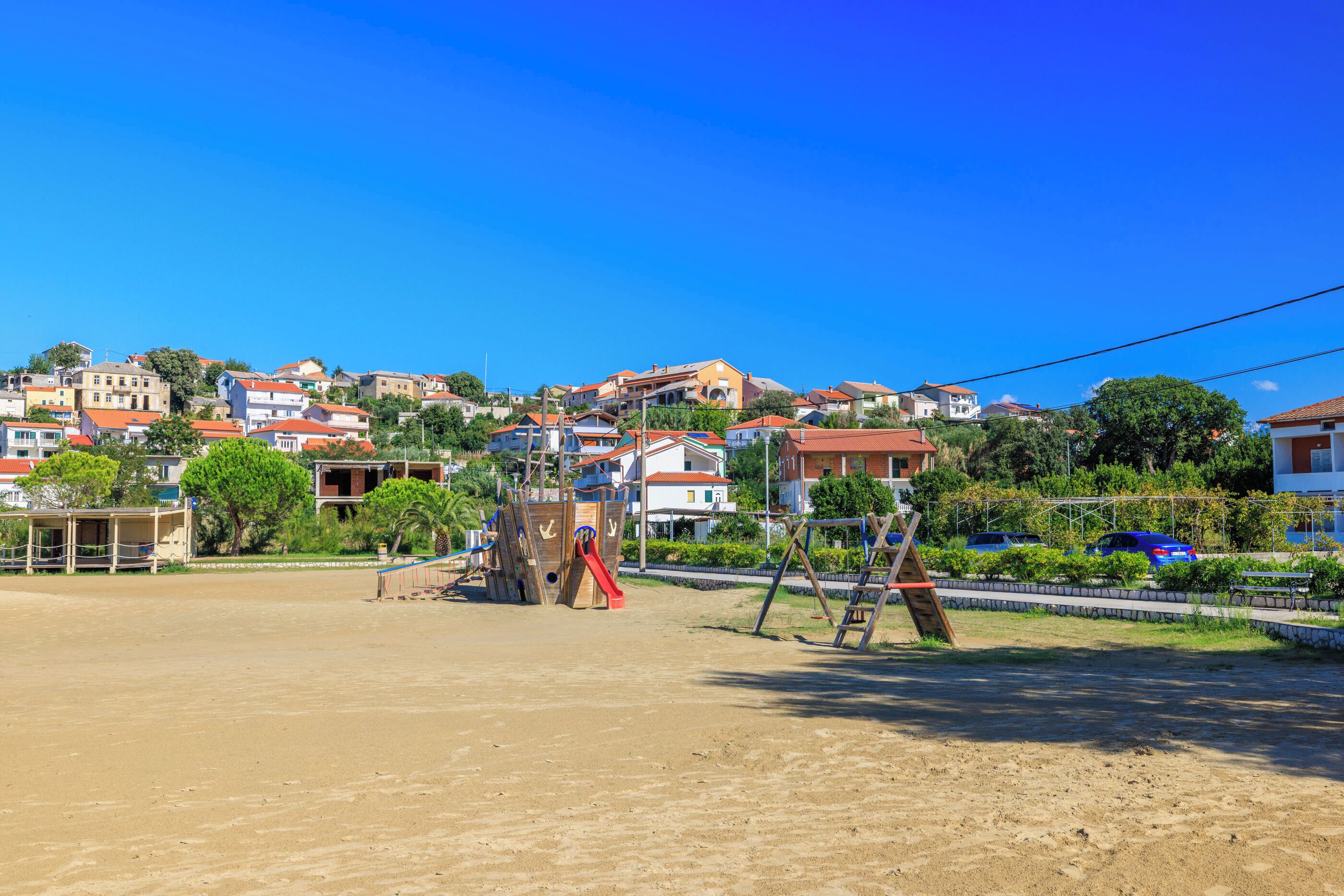 Área de recreação com areia, incluindo escorrega e baloiços, rodeada por edifícios residenciais.