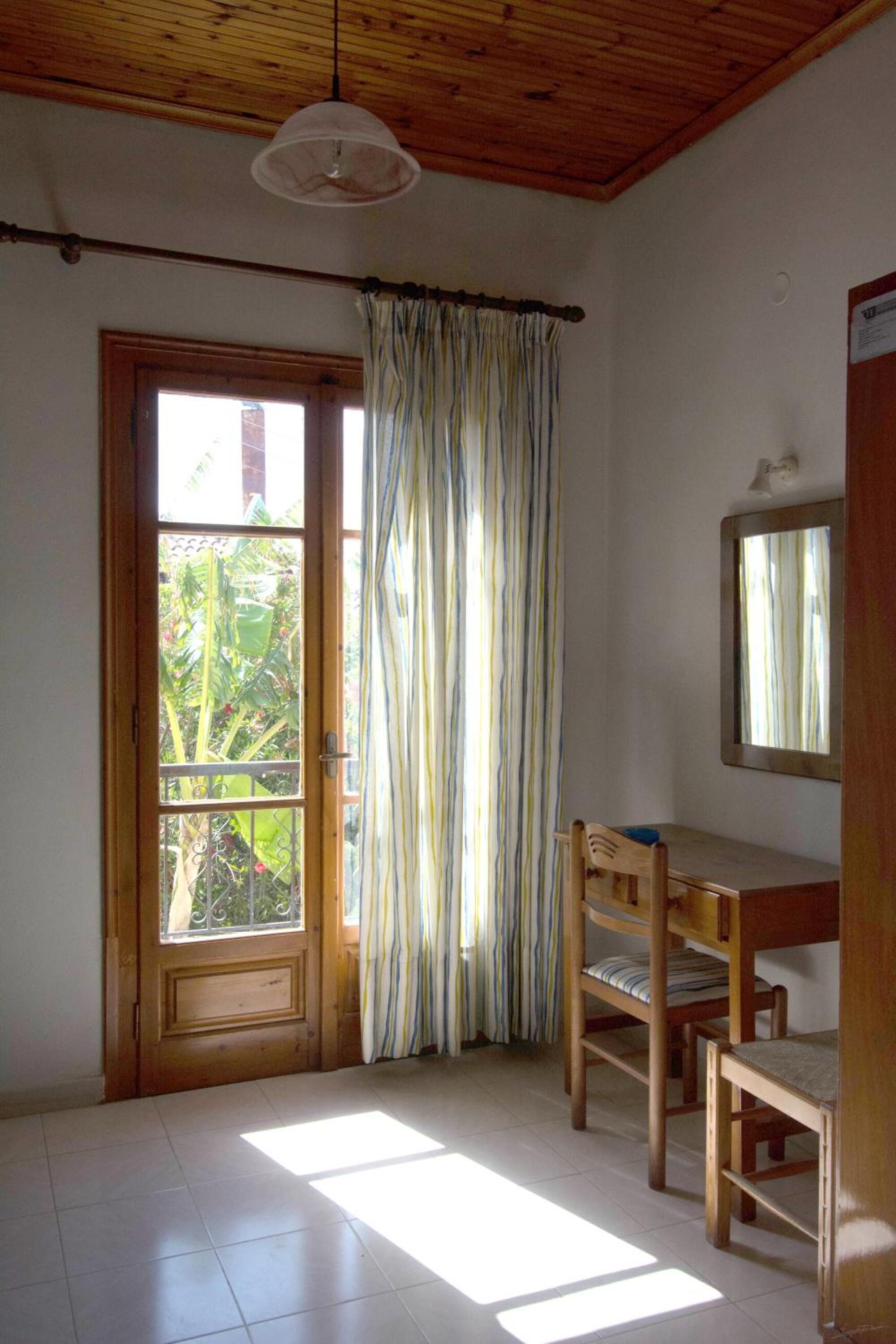 Quarto com porta de madeira, janela com cortinas, beliche, escrivaninha e espelho.