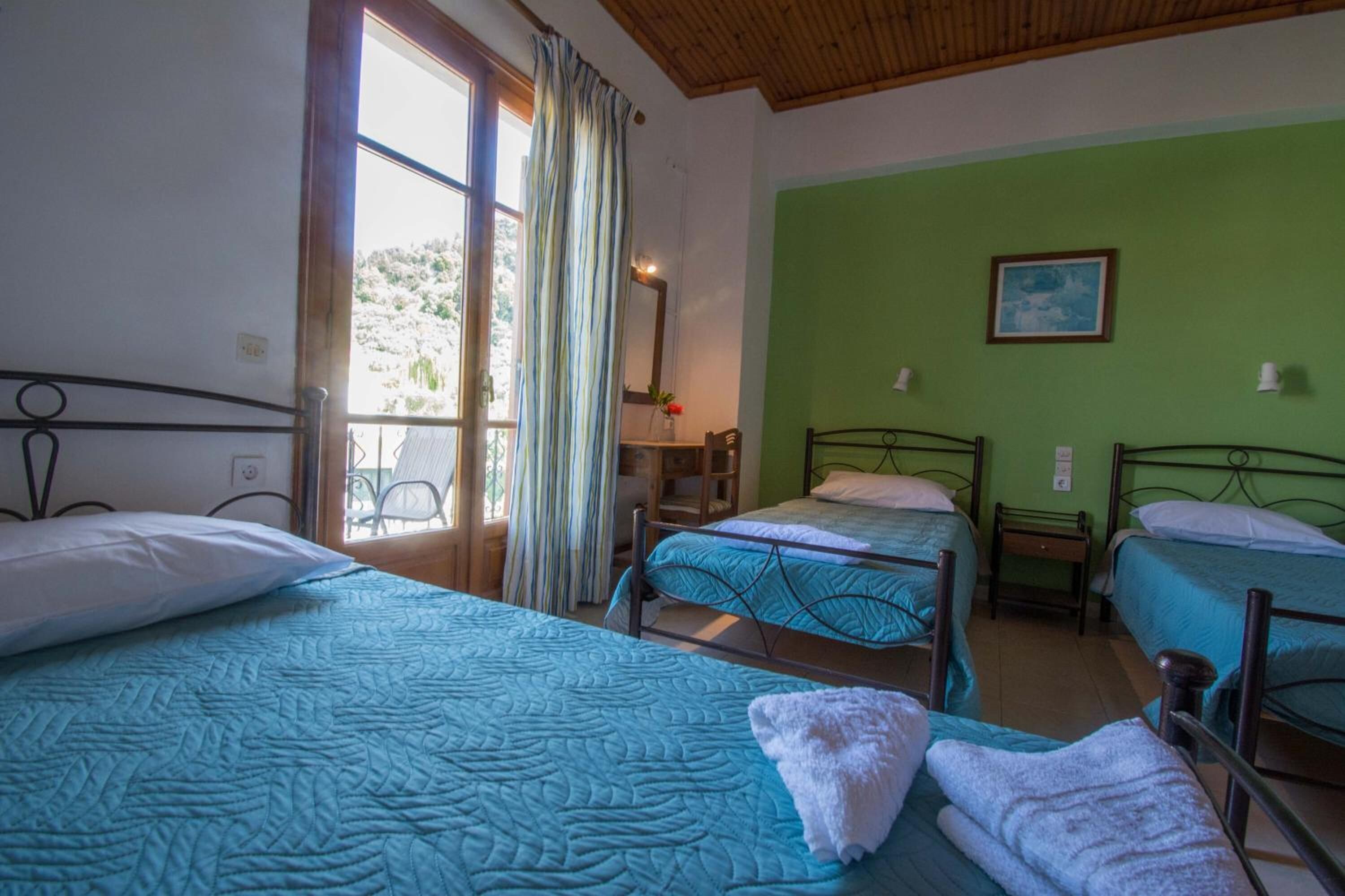Quarto com três camas, cada uma com colcha azul, cabeceira de madeira e travesseiro branco. Há uma janela com cortinas, uma pequena mesa com uma planta e um quadro na parede.