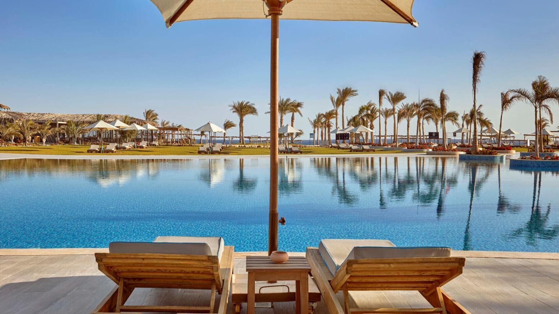 Área da piscina com espreguiçadeiras e guarda-sóis, cercada por palmeiras e edifícios do resort.