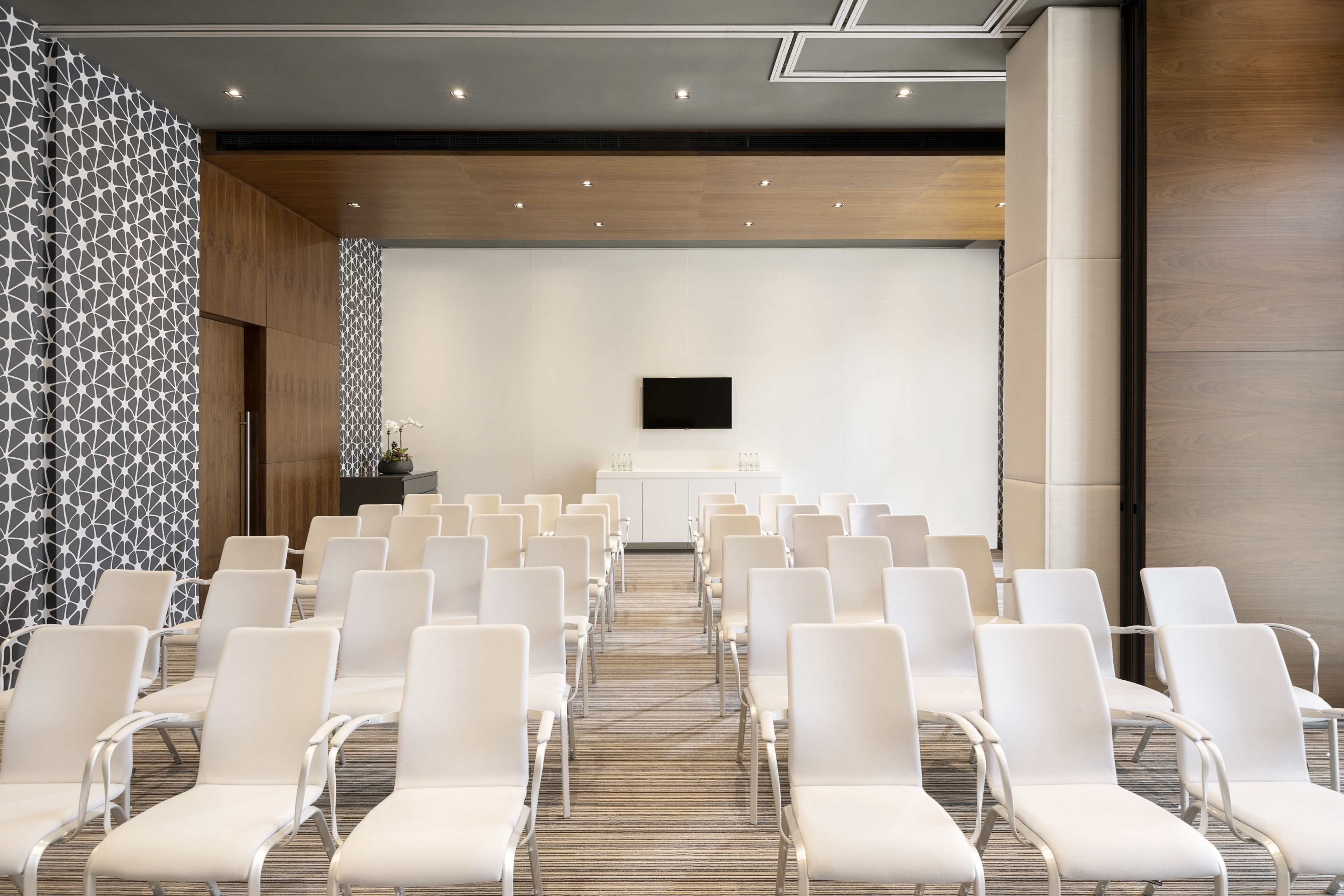 Uma sala de conferências com fileiras de cadeiras brancas, uma TV de tela plana e uma parede de painéis de madeira.